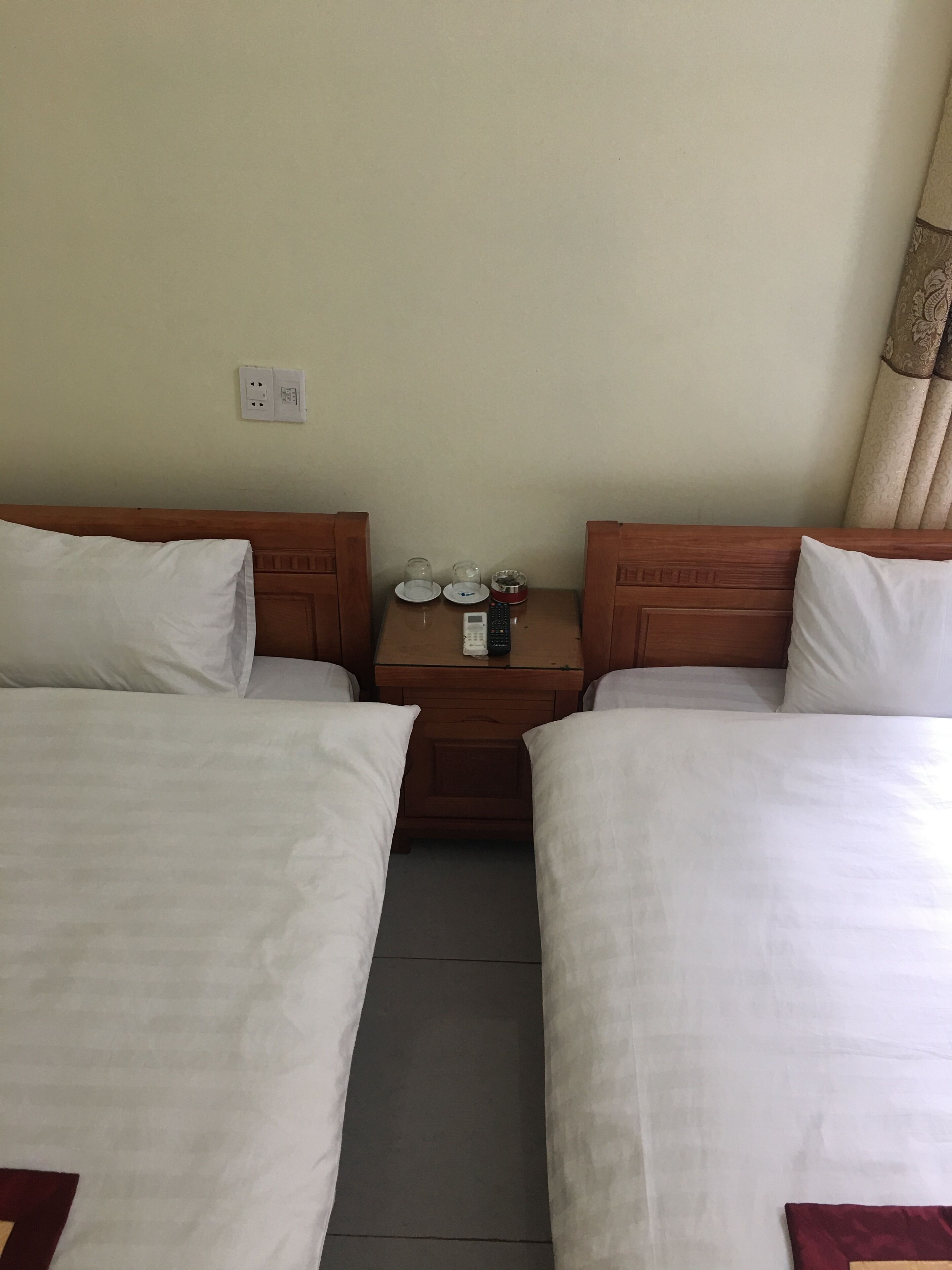 Quarto de hotel com duas camas de solteiro, mesa de cabeceira de madeira com um controle remoto, um copo e uma tigela.