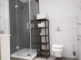 Uma casa de banho moderna com cabine de duche em vidro, sanita branca e prateleira de madeira com toalhas.