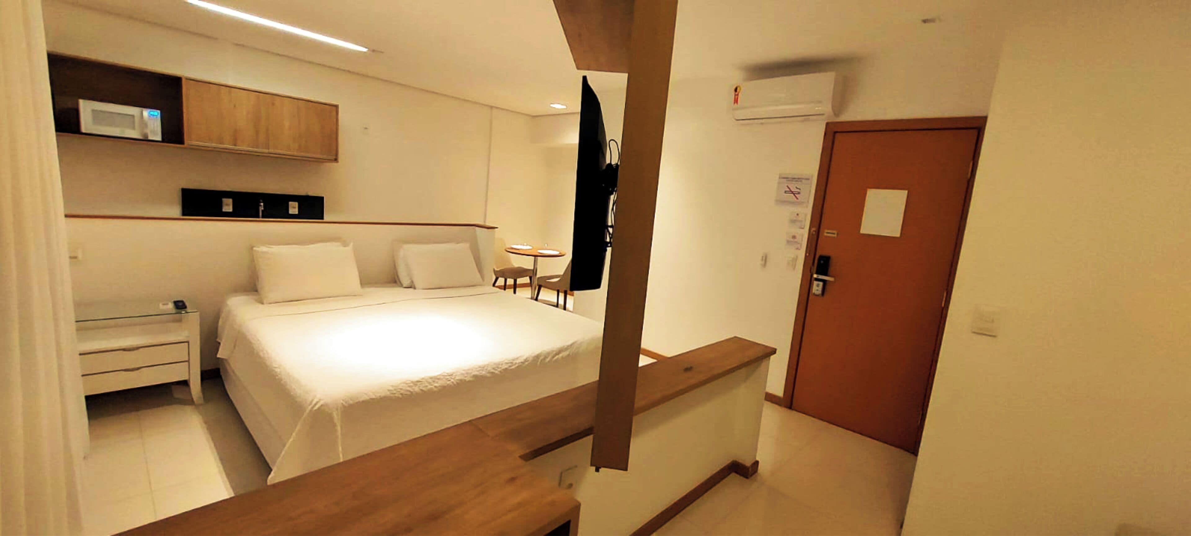 Quarto de hotel com cama, mesa de cabeceira, cadeira, mesinha, micro-ondas e porta com fechadura.