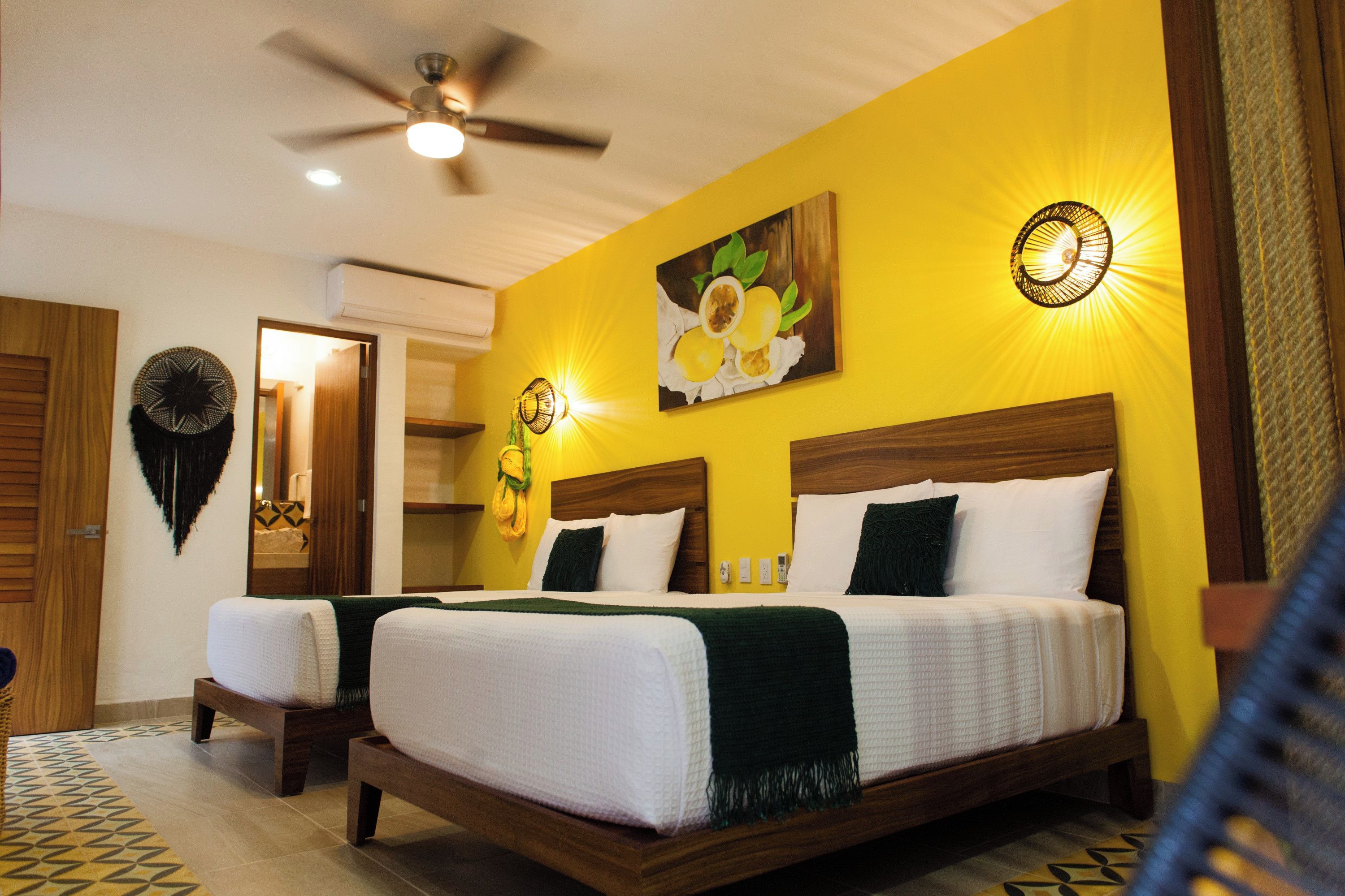 Quarto de hotel com duas camas, ventilador de teto, ar condicionado fixado na parede e uma pintura de frutas.