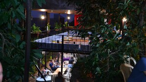 Uma área de refeições ao ar livre com mesas e cadeiras, rodeada por vegetação. Há uma ponte que liga dois níveis do restaurante.