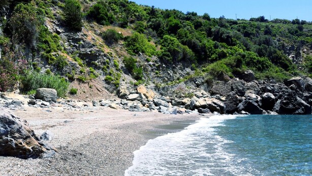 Uma praia rochosa com águas azul-claras, rodeada por vegetação exuberante e colinas.