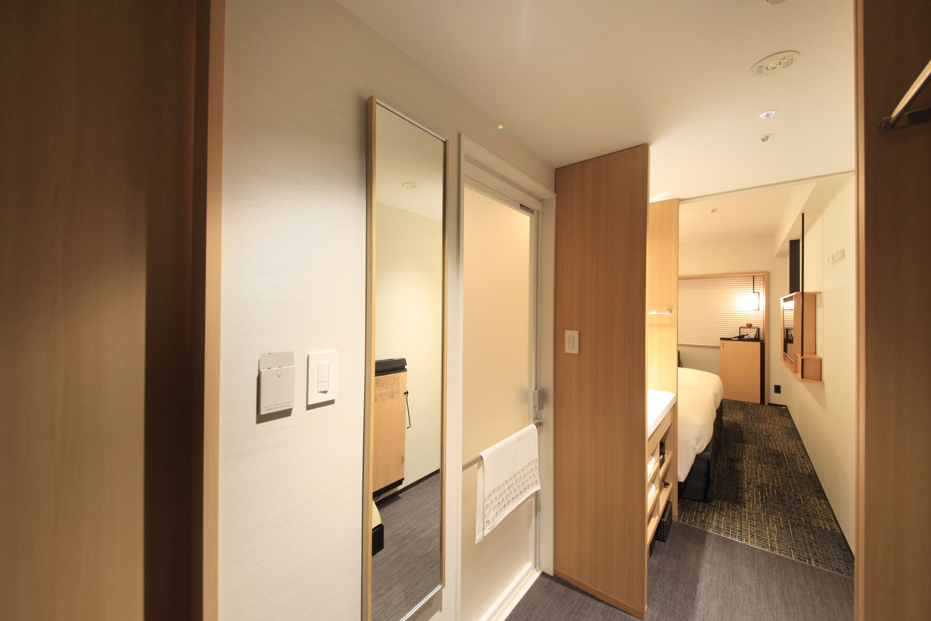 Corredor de hotel com uma cama, uma escrivaninha pequena e um espelho.