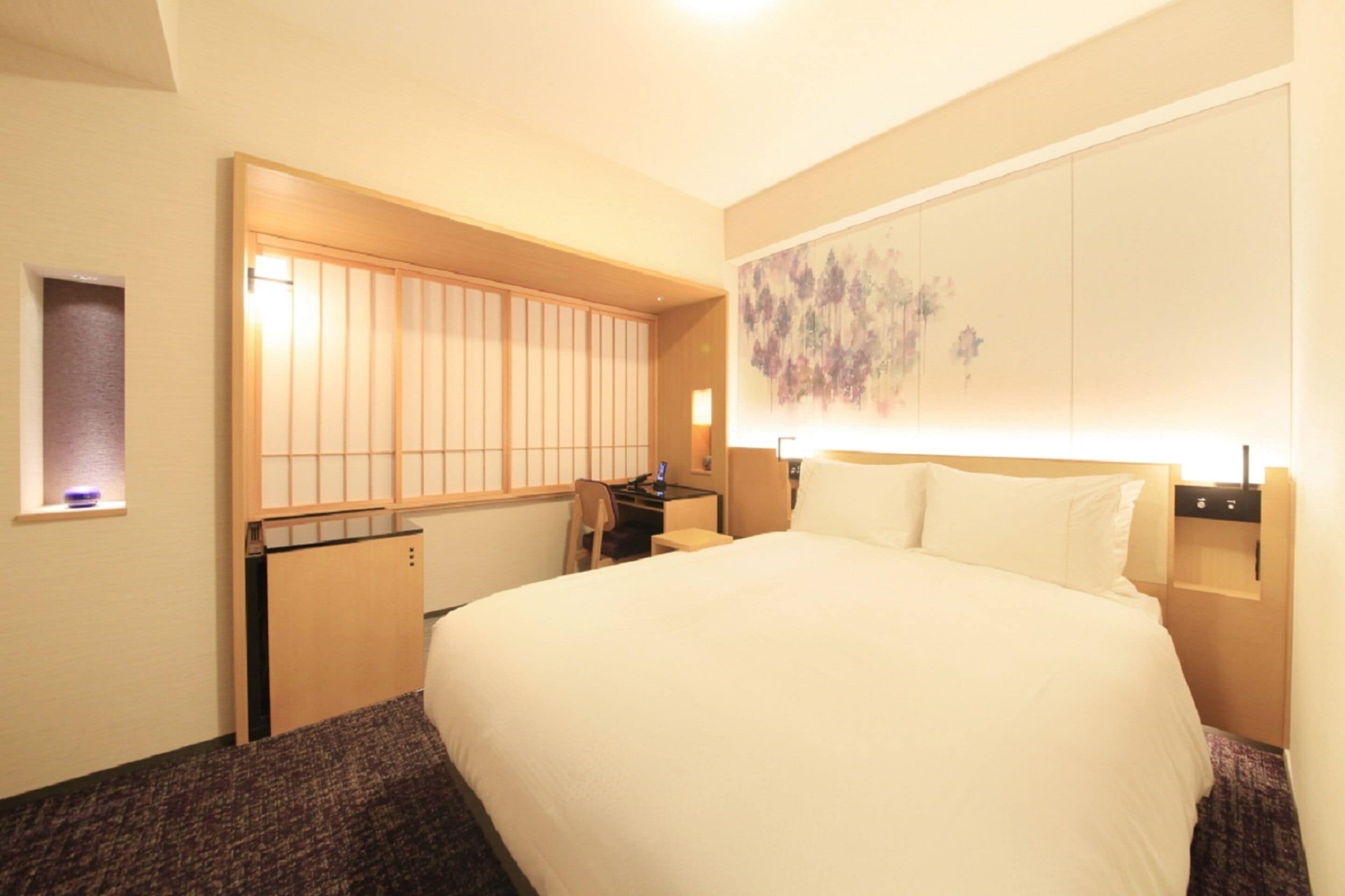 Quarto de hotel com uma cama grande, uma mesa e uma cadeira. Há uma televisão na parede e um quadro sobre a cama.