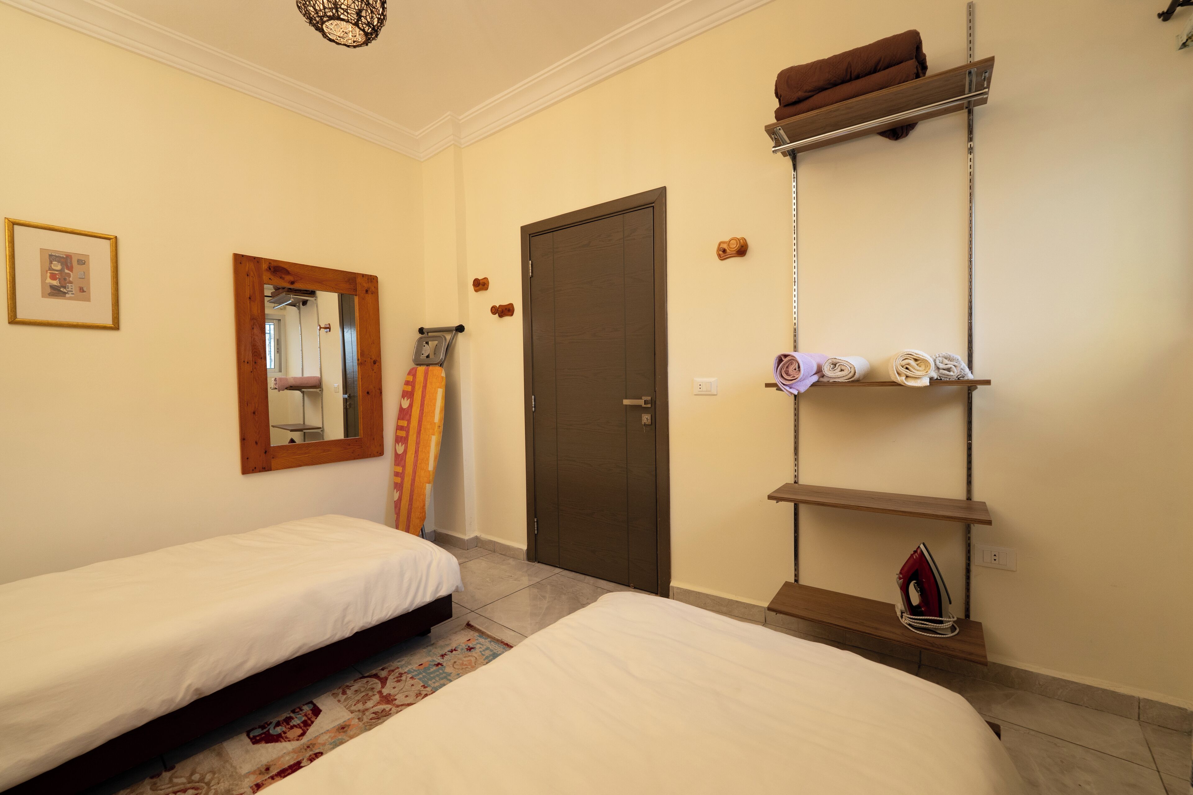 Quarto com duas camas de solteiro, uma prateleira de madeira com toalhas, uma porta e um espelho.