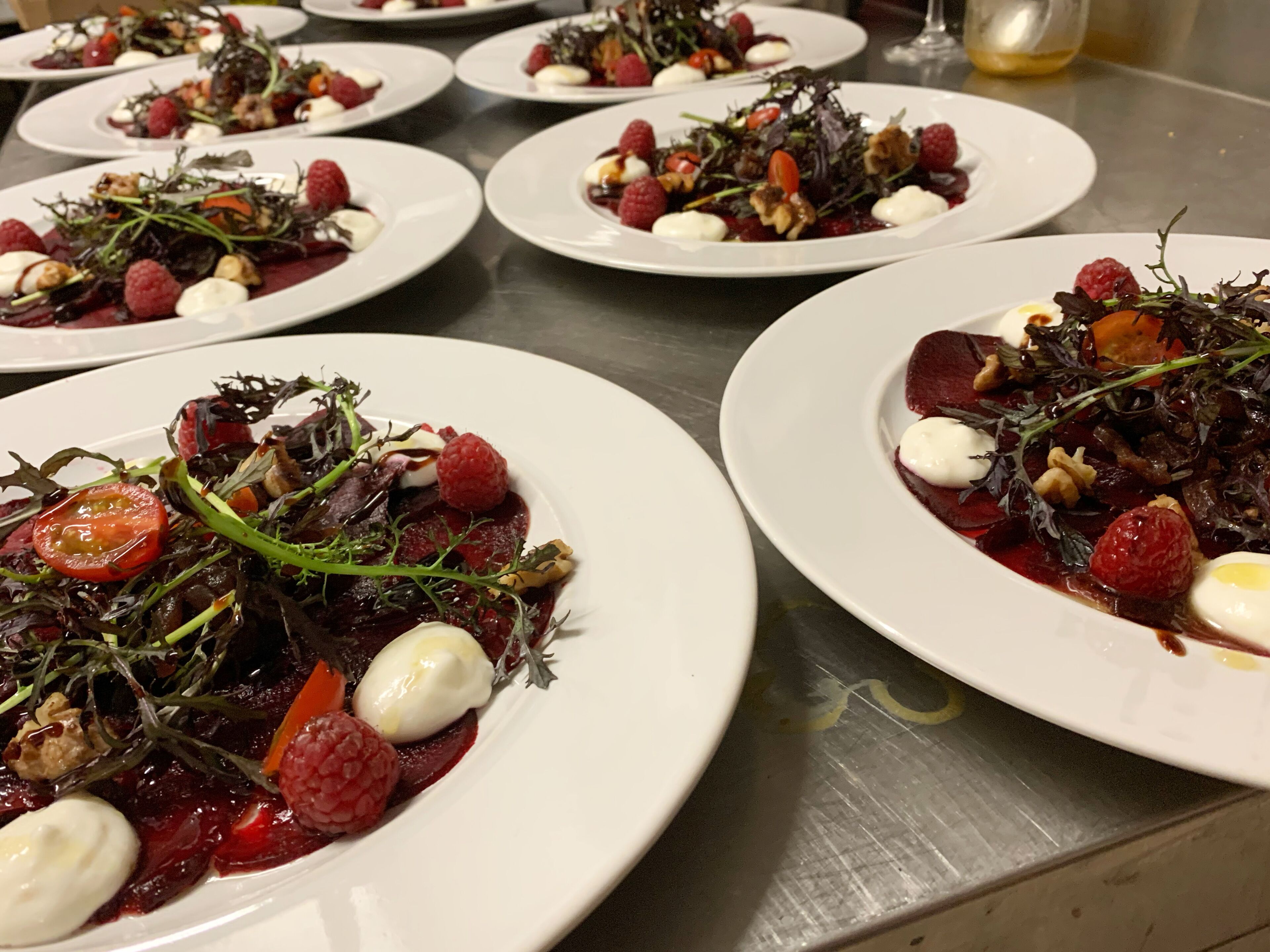Uma série de pratos brancos com uma salada de folhas verdes, framboesas e um creme branco, dispostos sobre uma superfície de aço inoxidável.