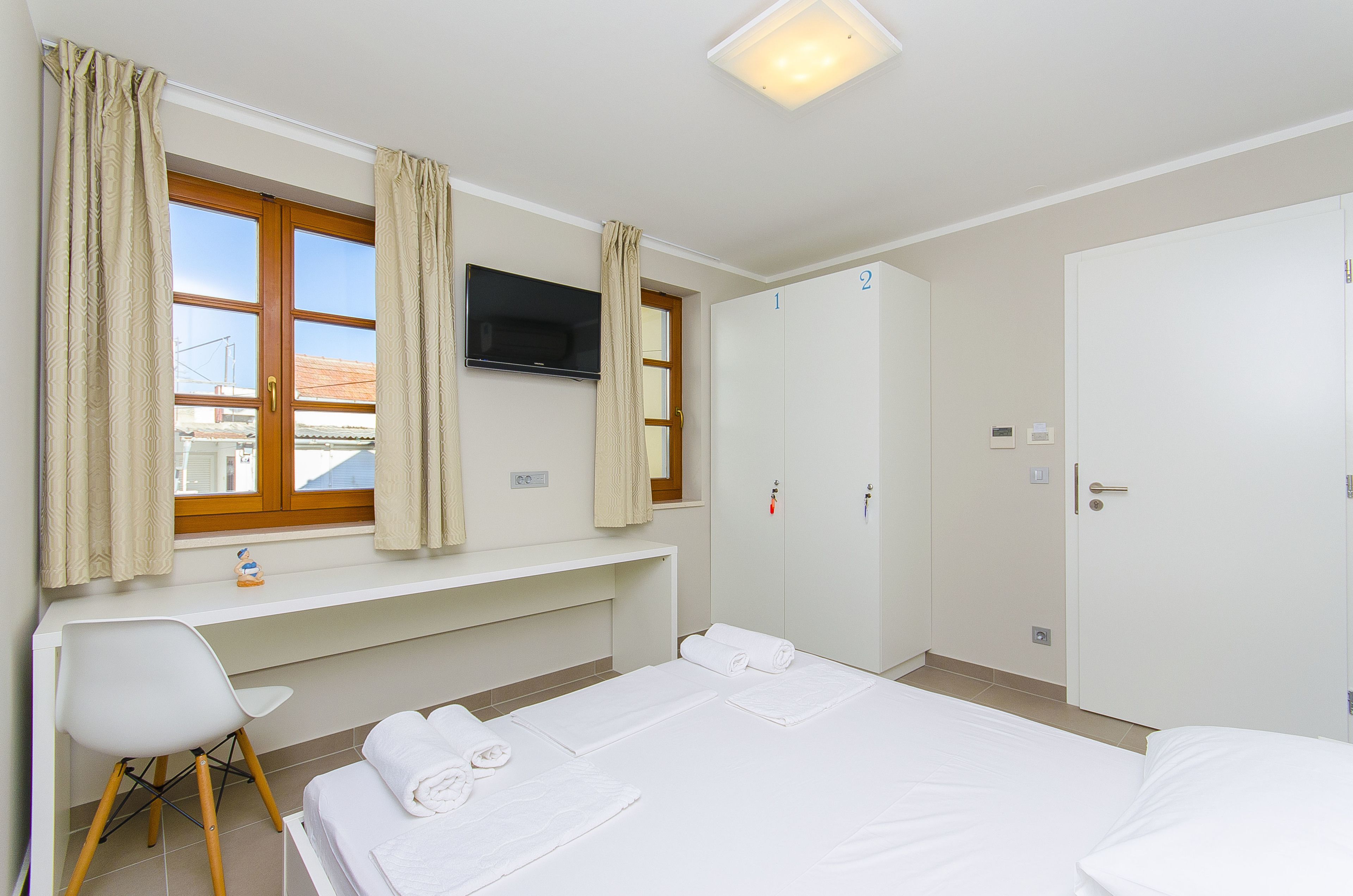 Quarto de hotel com cama, escrivaninha, cadeira, televisão e guarda-roupa.