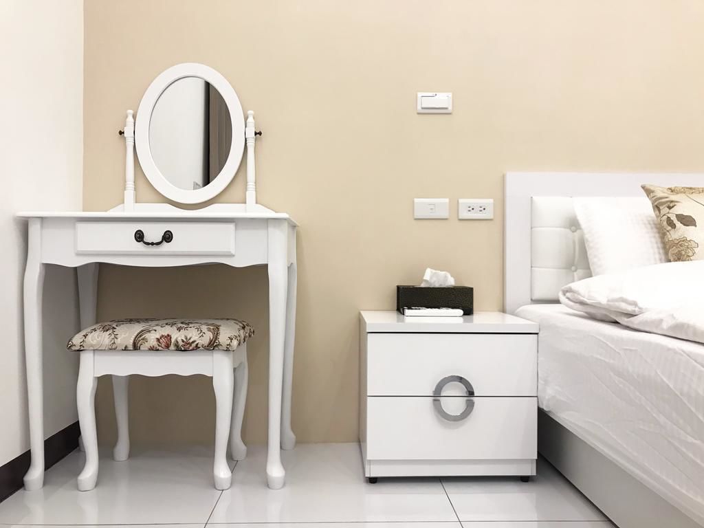 Quarto com uma cama branca, uma mesinha de cabeceira com um estojo de lenços, cômoda branca com espelho e uma poltrona floral, e parede bege.