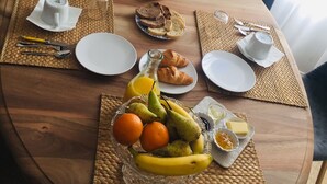 Mesa de café da manhã posta com pratos, talheres, garrafa de água, torradeira e diversos alimentos, incluindo pão, queijo, frutas e doces.
