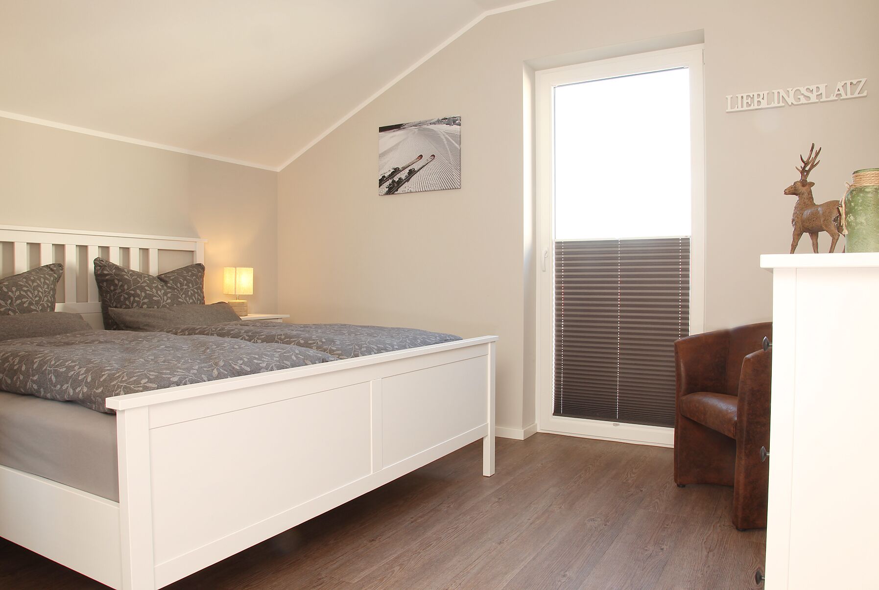 Quarto com cama branca, mesa de cabeceira com luminária, janela com persianas e prateleira na parede com objetos decorativos.