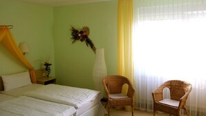 Quarto de hotel com duas camas, uma cadeira de vime, uma mesa de cabeceira com abajur e um objeto decorativo na parede.