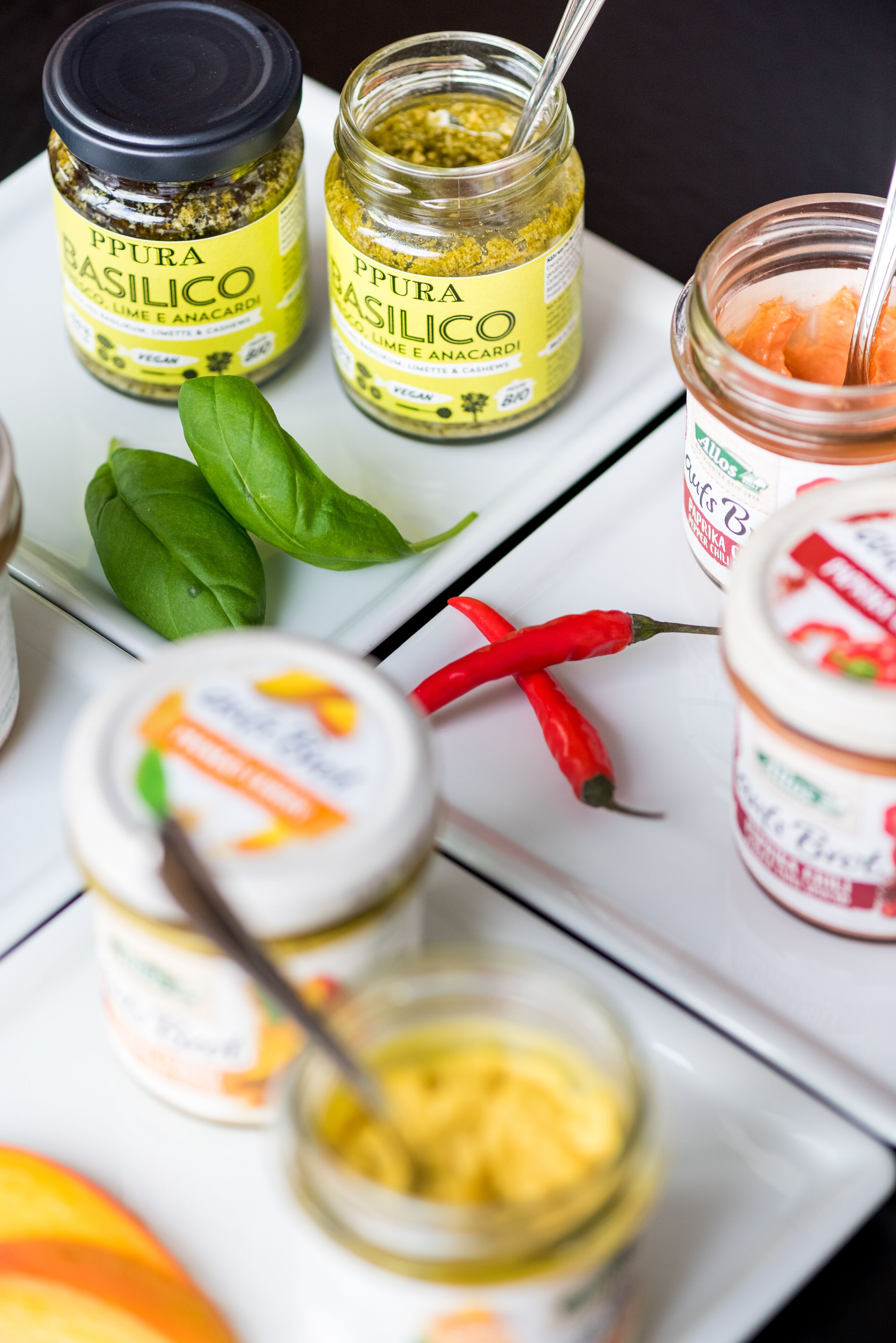 Uma mesa com diversos alimentos, incluindo potes de pesto, uma tigela de pasta amarela e folhas de manjericão fresco.
