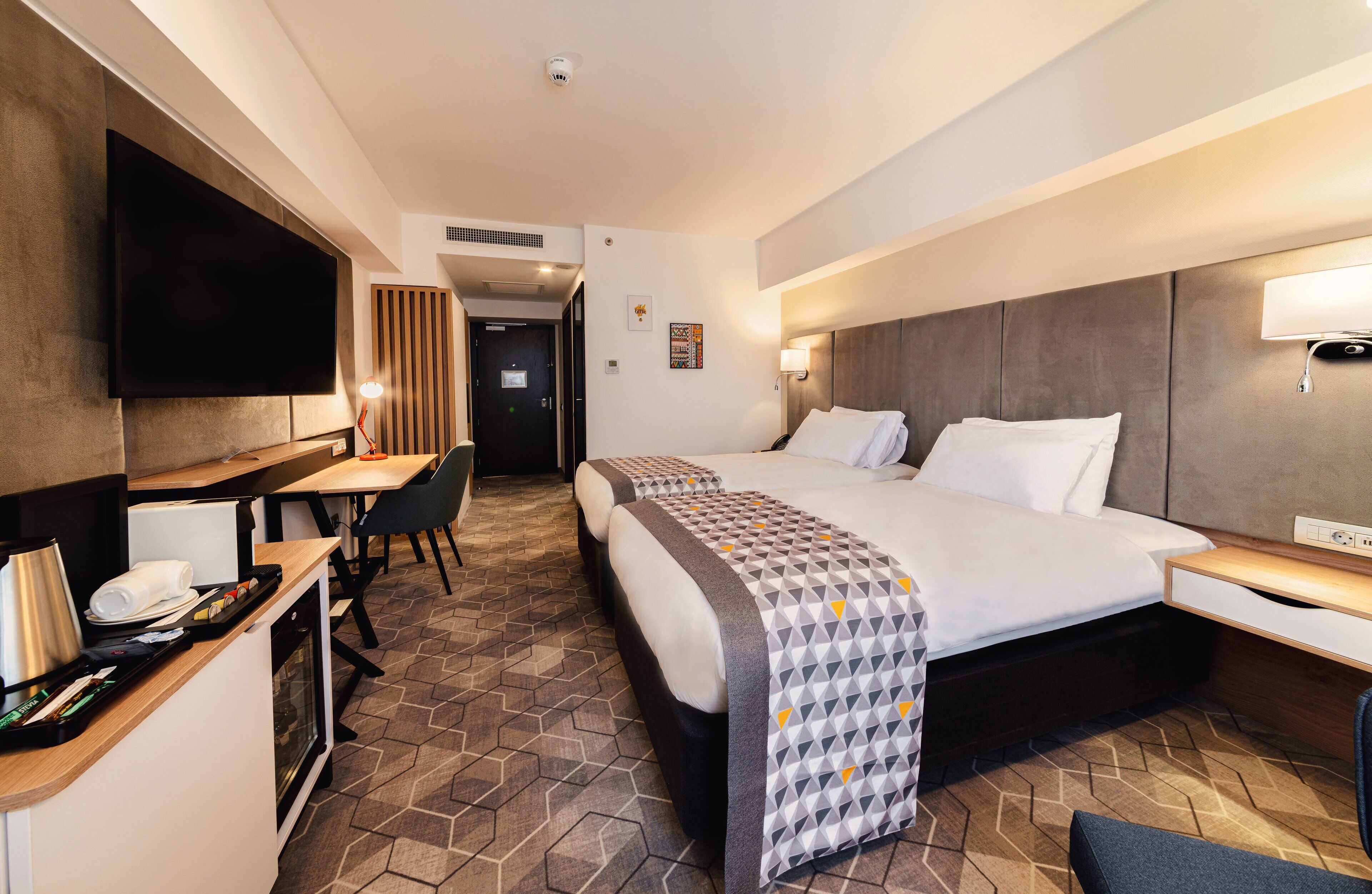 Quarto de hotel com uma cama grande, uma mesa com televisão de tela plana, uma cozinha compacta e uma área de estar separada.