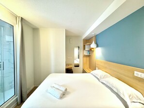 Quarto de hotel com uma cama grande, janela com cortinas, cabeceira de madeira e mesa de cabeceira com abajur.