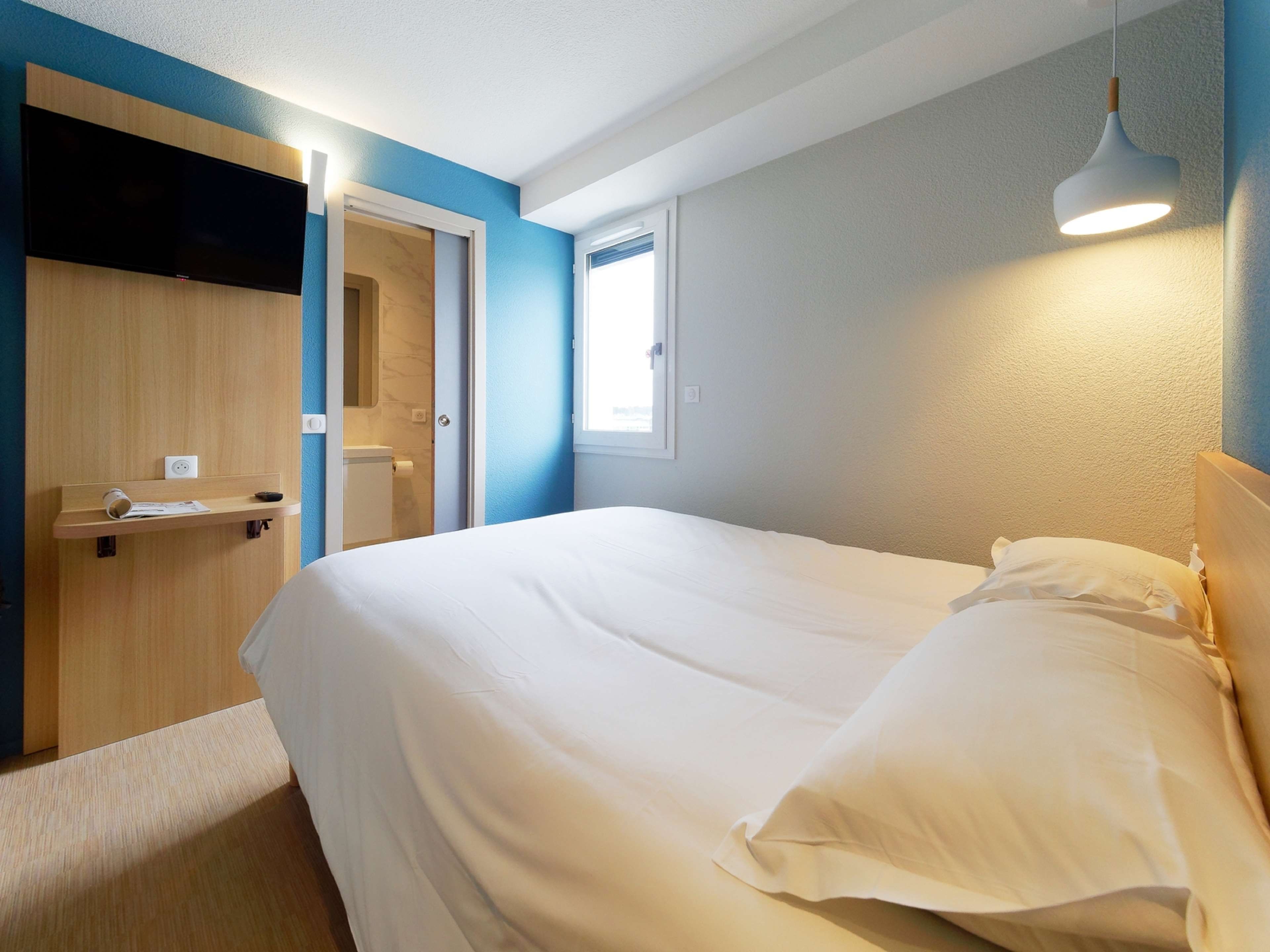 Quarto de hotel com cama, prateleira de madeira, televisão e janela.
