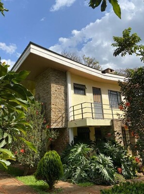Uma casa de dois andares com fachada de pedra, varanda com grade de metal e um jardim com diversas plantas e flores.