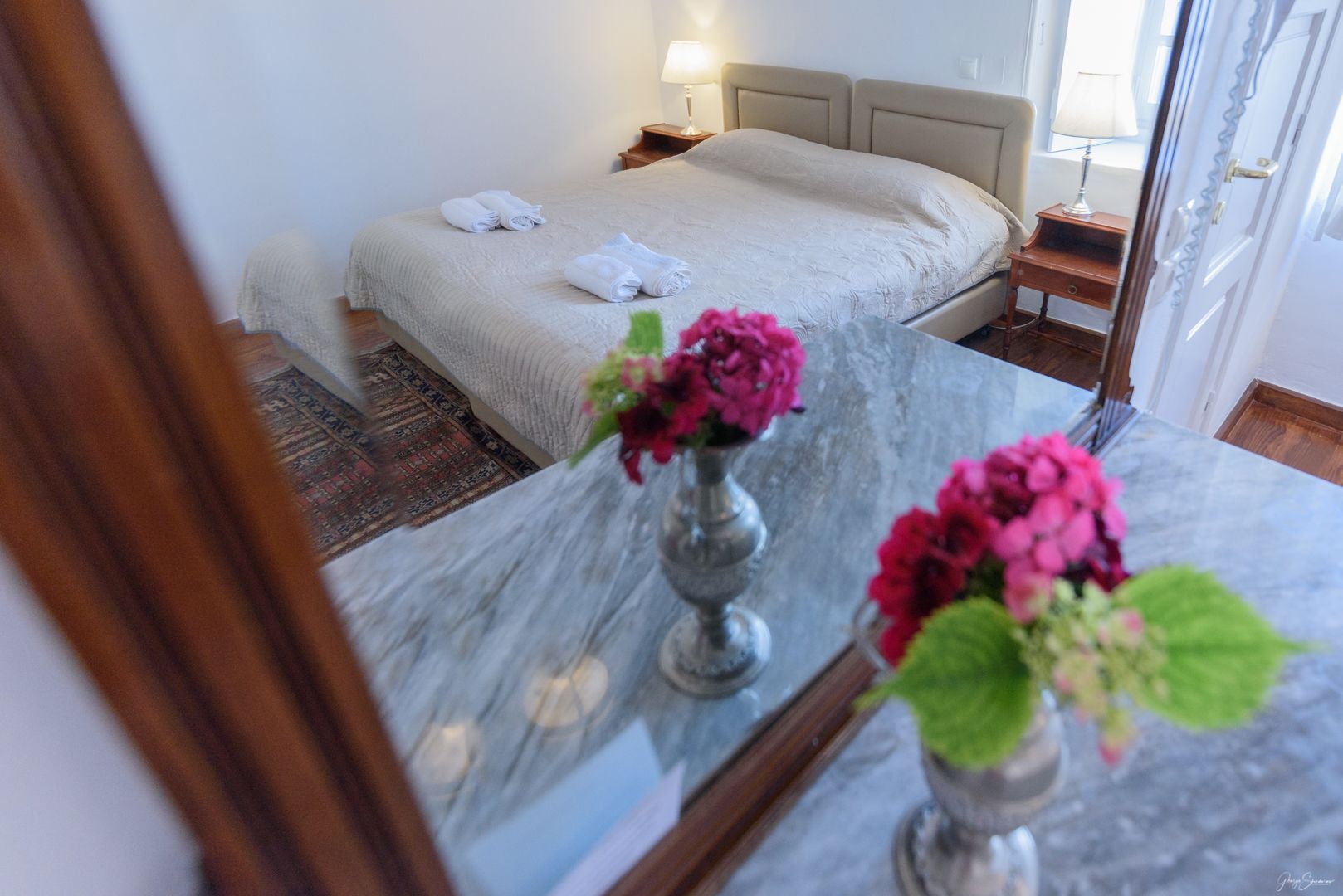 Quarto de hotel com cama, mesas de cabeceira, espelho e um vaso com flores.