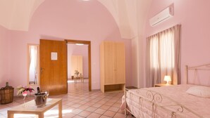 Quarto com cama, guarda-roupa de madeira, mesa com garrafa e copos, e porta que dá para outro espaço.