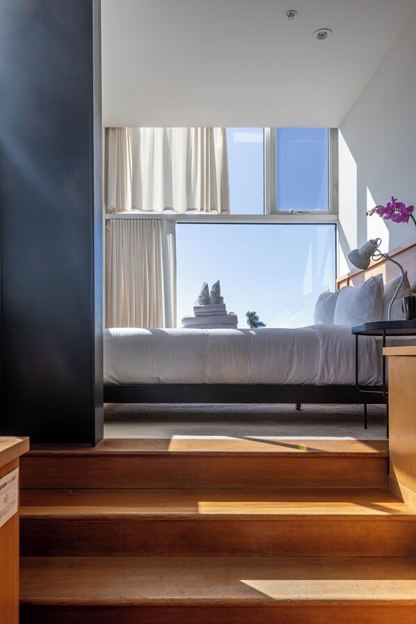 Quarto com cama, mesa de cabeceira, abajur e janela com cortinas.