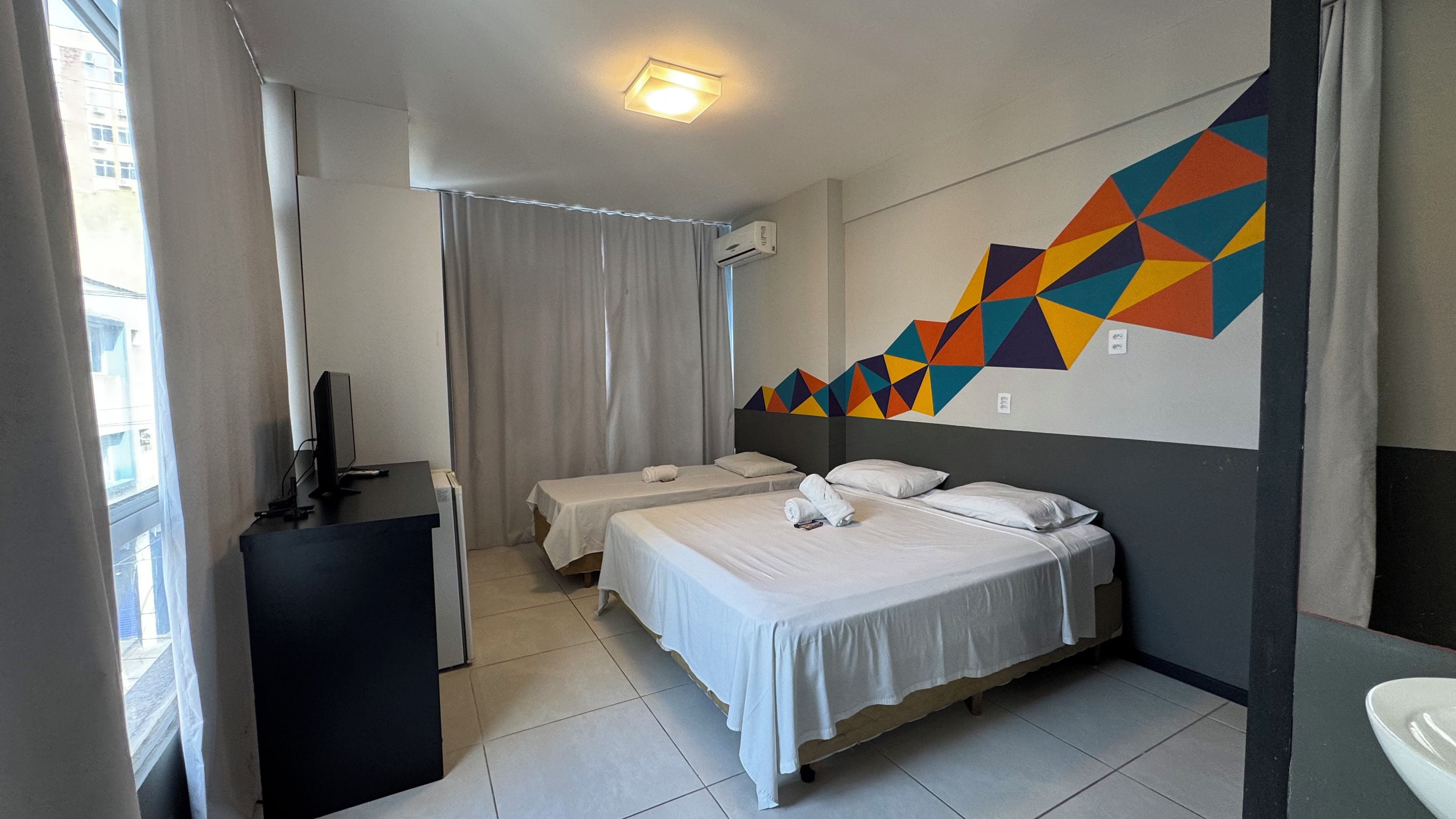 Quarto de hotel com duas camas, uma escrivaninha, uma televisão e uma decoração geométrica na parede.