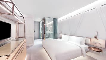 Quarto de hotel moderno com cama grande, televisão, casa de banho com paredes de vidro e secretária embutida.