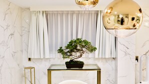 Um interior moderno com parede de mármore, uma árvore bonsai sobre mesa de vidro e um espelho com moldura dourada.