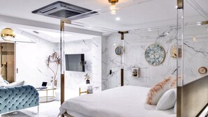 Quarto moderno com uma cama grande, uma poltrona azul estofada, uma pequena escrivaninha com um laptop e uma TV fixada na parede.