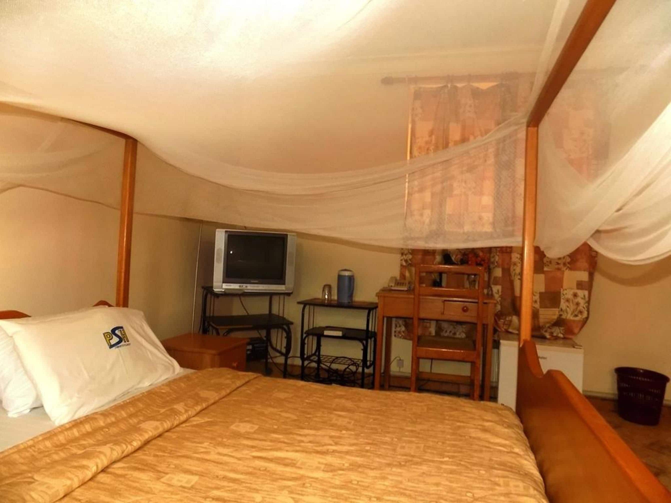 Uma cama de solteiro com dossel, uma televisão sobre suporte, uma mesinha e uma cadeira.