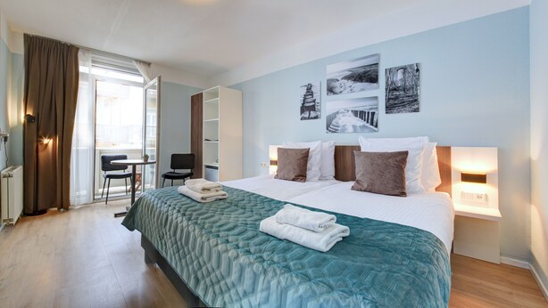 Quarto de hotel com uma cama grande, uma escrivaninha e uma cadeira. Há três luminárias fixadas na parede e uma janela com cortinas.