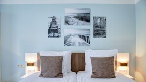 Uma cama de casal com almofadas brancas e castanhas, uma cabeceira de madeira e luminárias fixadas na parede.