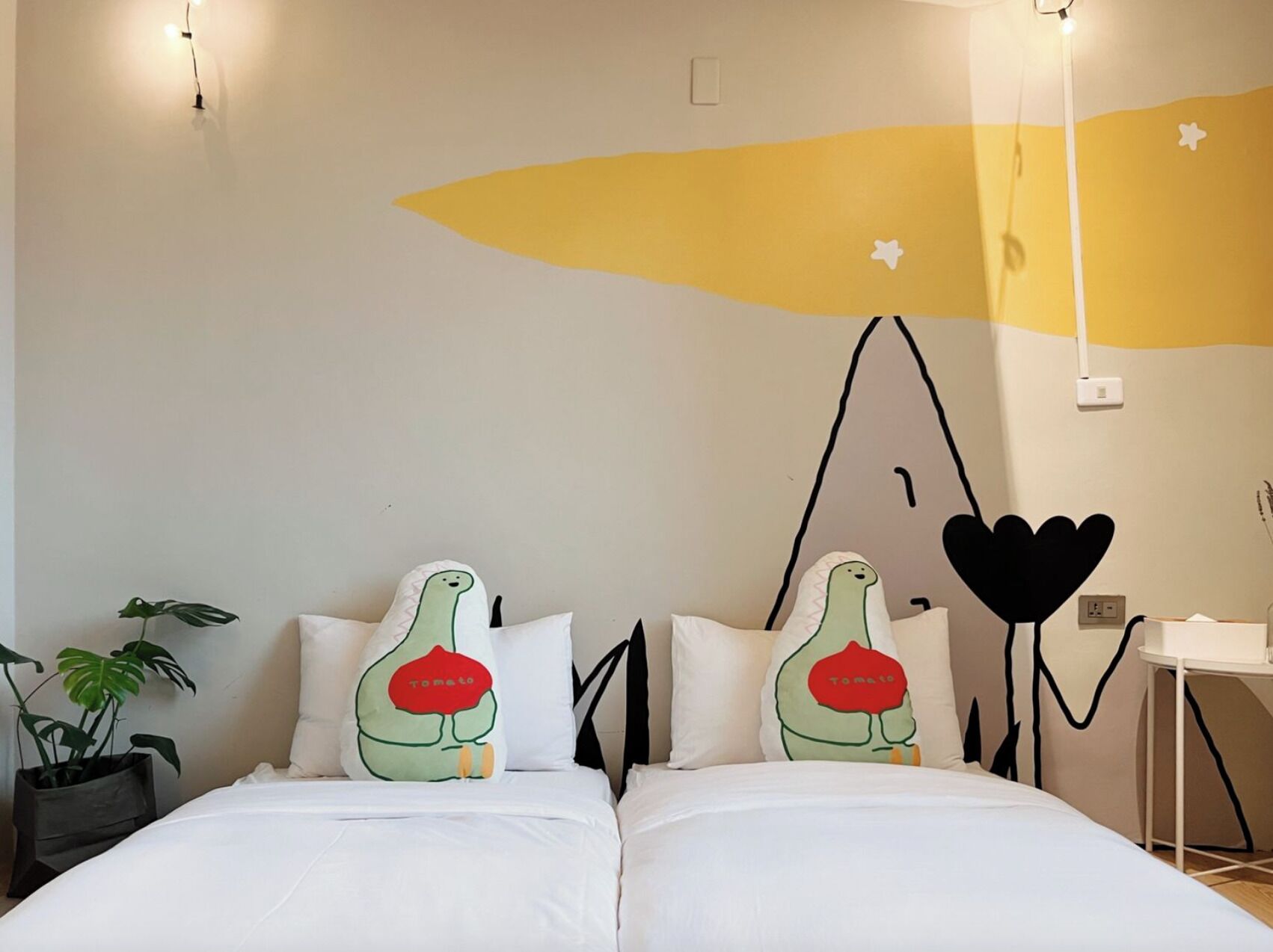 Quarto com duas camas, um mural na parede e uma pequena mesa com uma planta.