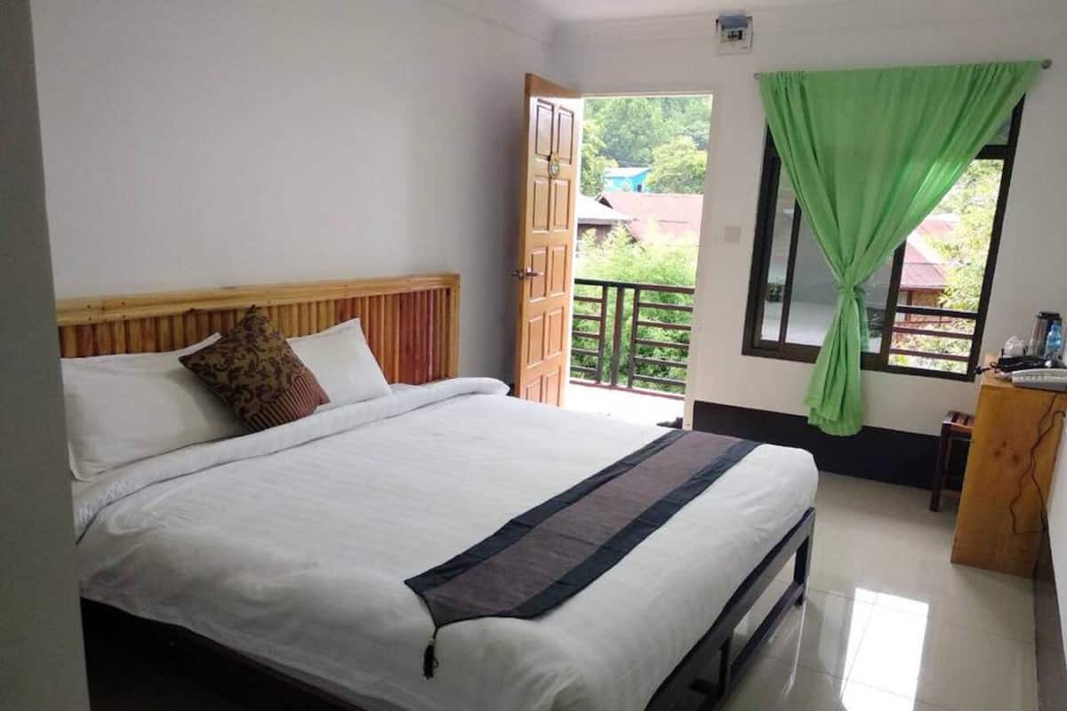 Quarto com cama, cabeceira de madeira, cortina verde e janela com vista para vegetação.