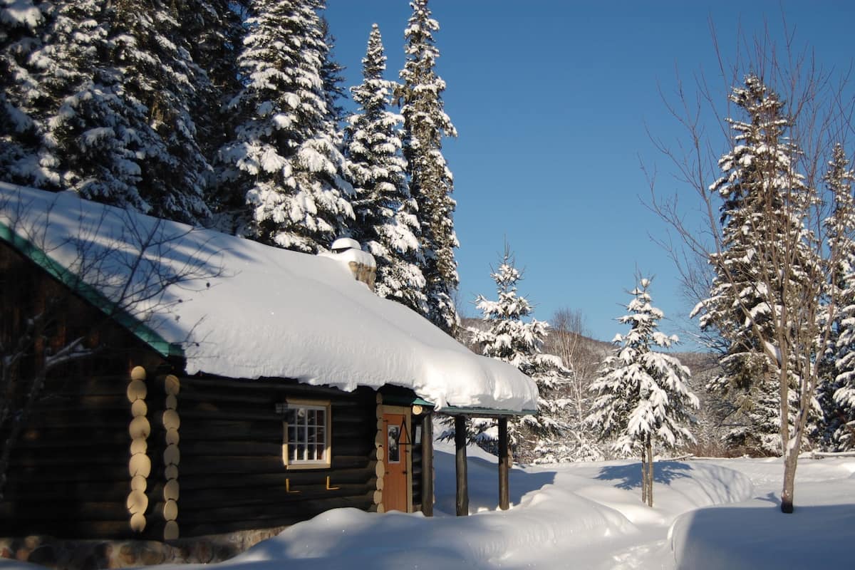 Uma cabana de madeira com telhado coberto de neve, rodeada por árvores carregadas de neve.