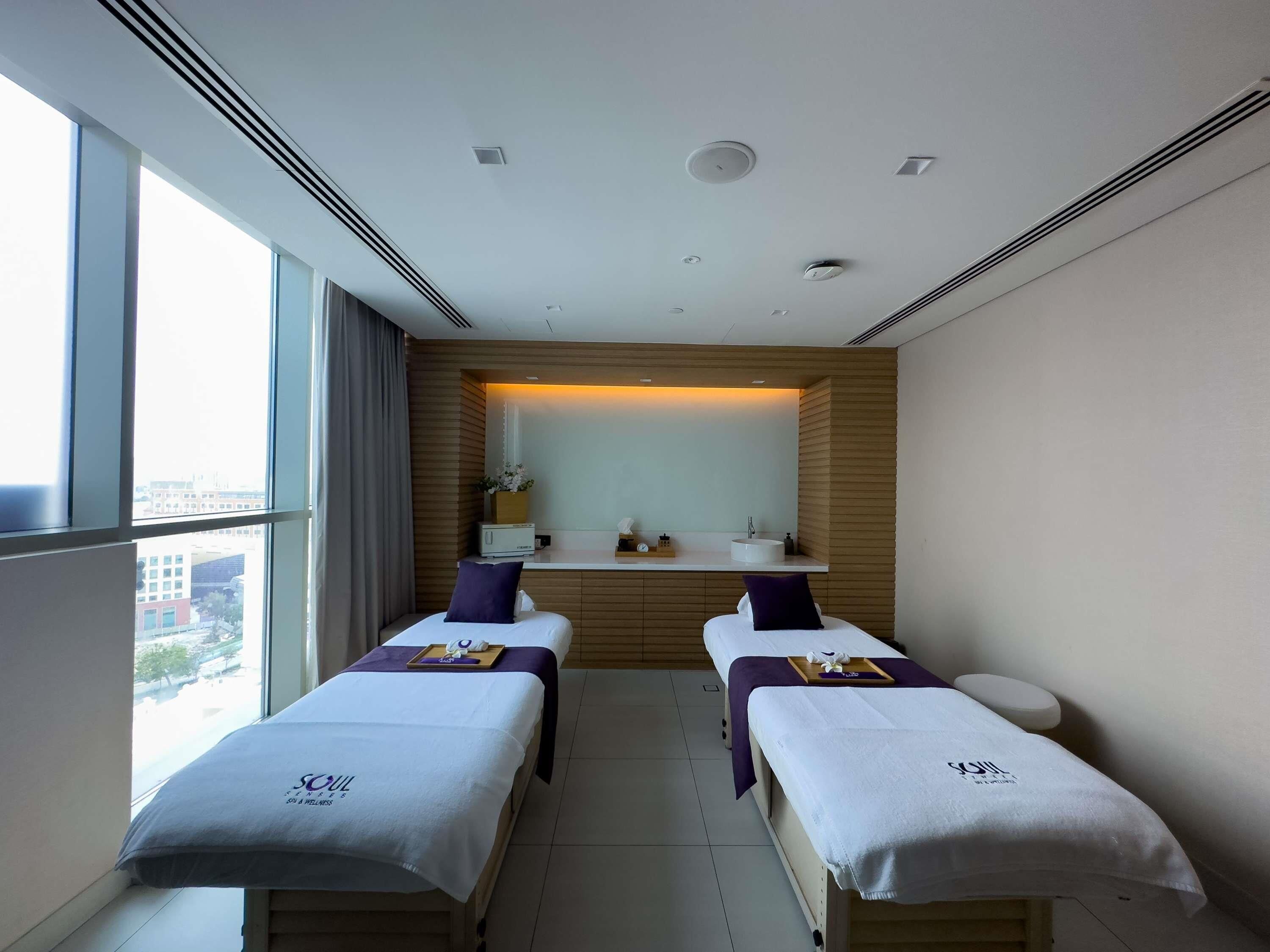 Quarto de hotel moderno com duas macas de massagem, uma pequena toucador com pia e uma grande janela com vista para a cidade.