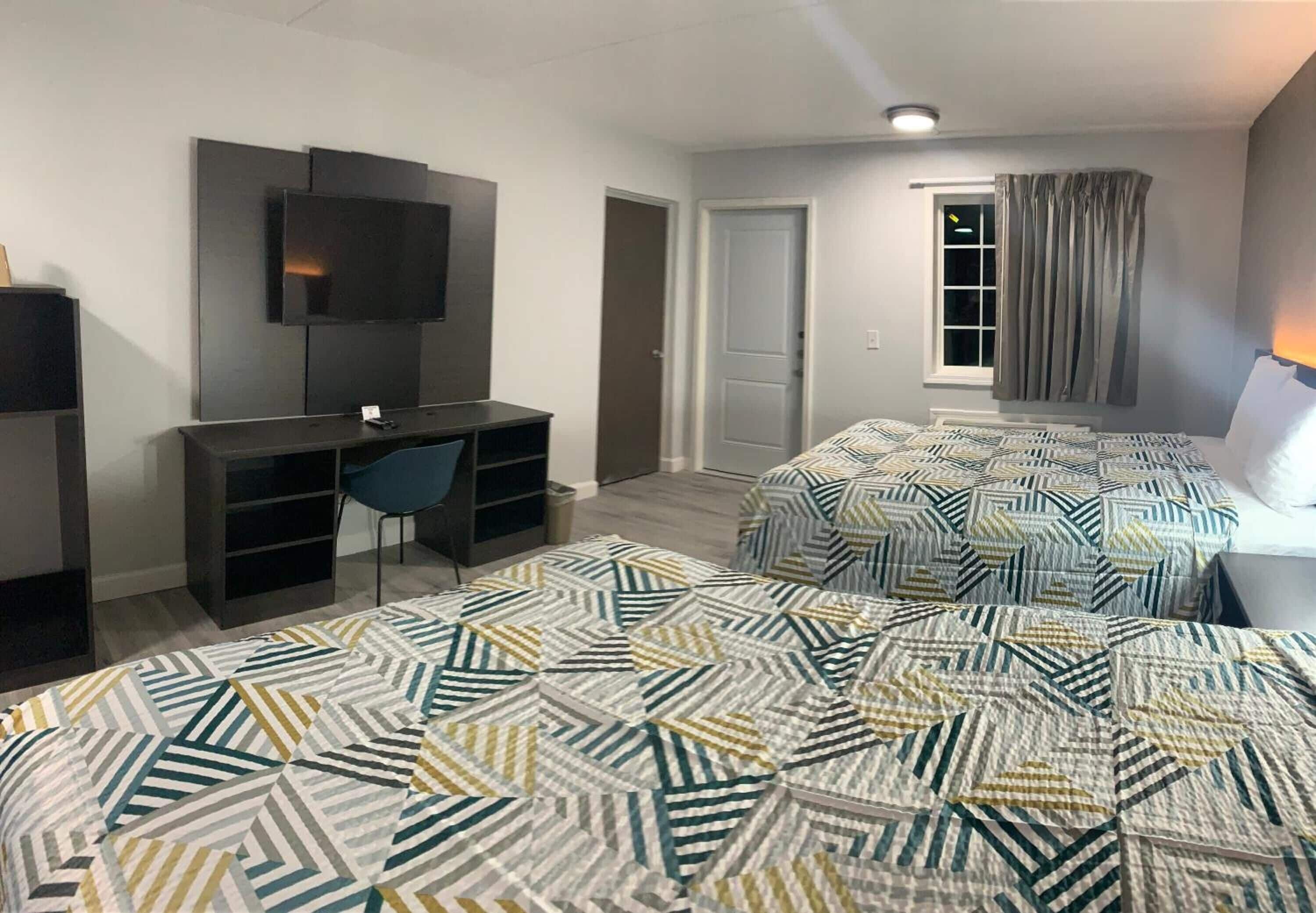 Quarto com cama, escrivaninha, cadeira, televisão e janela com cortinas.