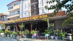 Um hotel chamado 'Neoss Tel' com um café chamado 'Resto Cafe Bar' em frente. Há plantas em vasos e uma bicicleta estacionada lá fora.