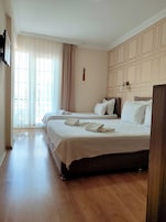 Quarto de hotel com duas camas, piso de madeira, uma janela grande com cortinas e televisão presa na parede.