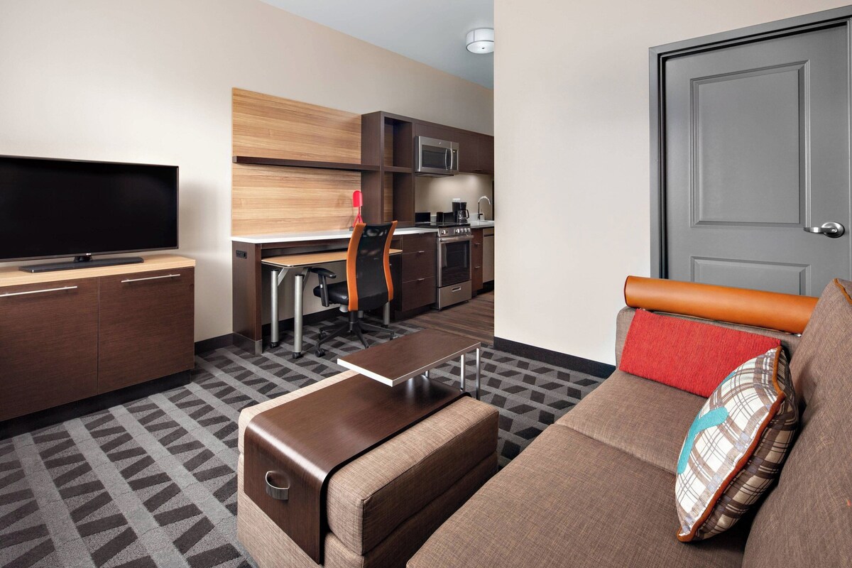 Quarto de hotel compacto com cozinha compacta, televisão de tela plana, sofá com almofada vermelha e uma porta.