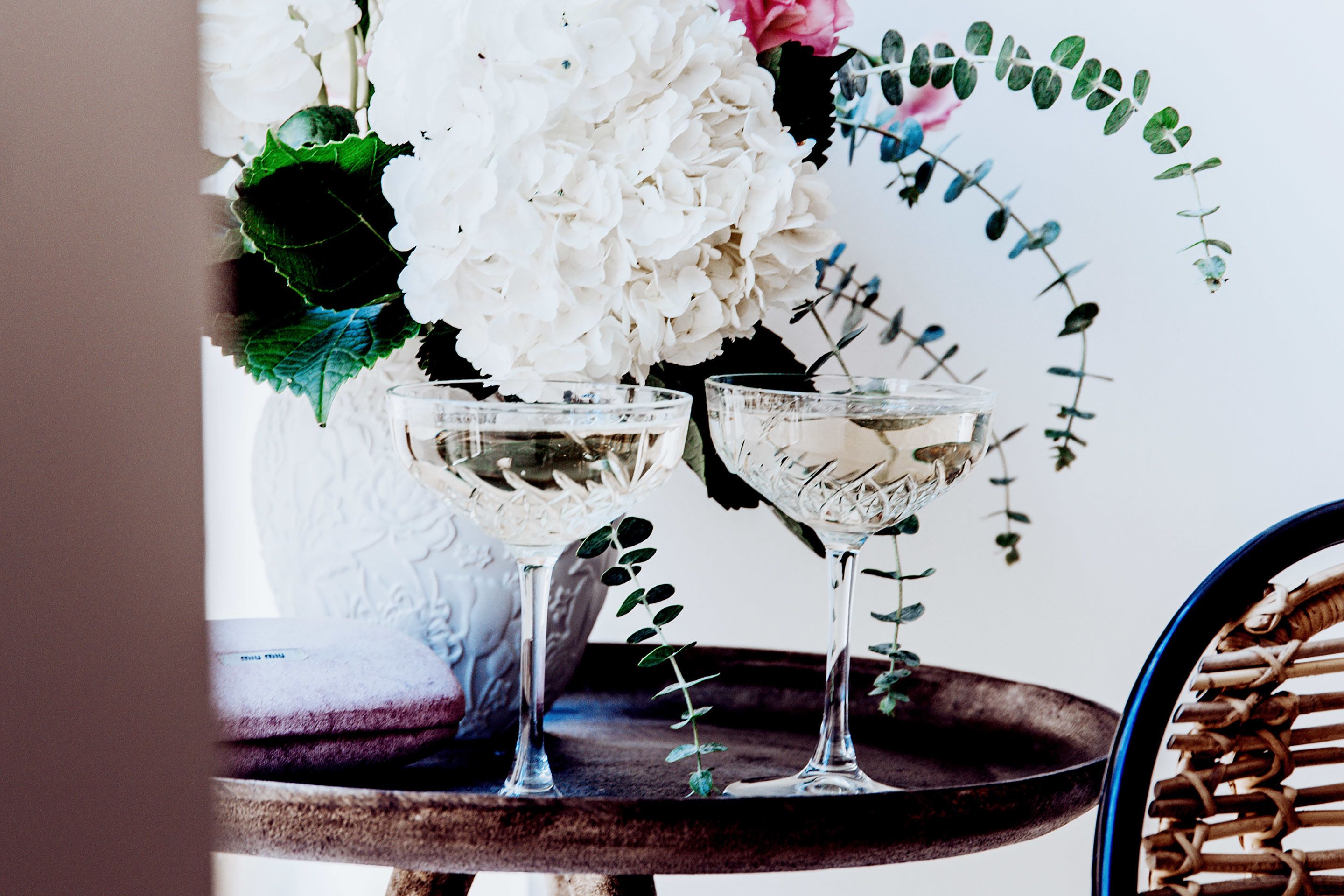 Uma mesa com dois copos de vidro transparente, um vaso branco com flores e uma cadeira de vime.