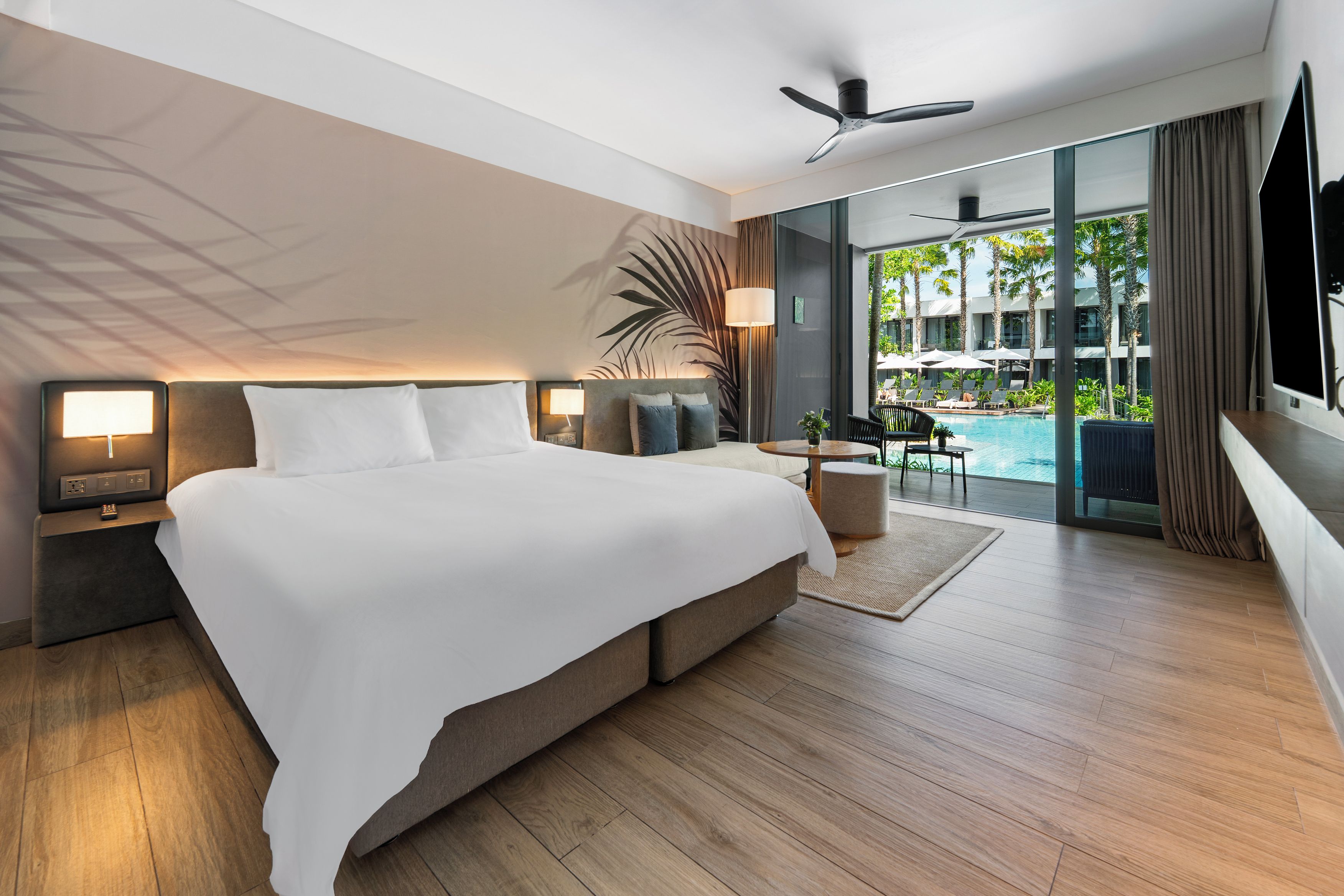 Quarto de hotel moderno com uma cama grande, ventoinha no teto, TV e vista para uma piscina e área de estar externa.