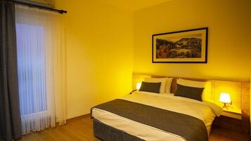 Quarto com cama, duas luminárias de cabeceira, mesa de cabeceira, janela com cortinas e quadro pendurado na parede.
