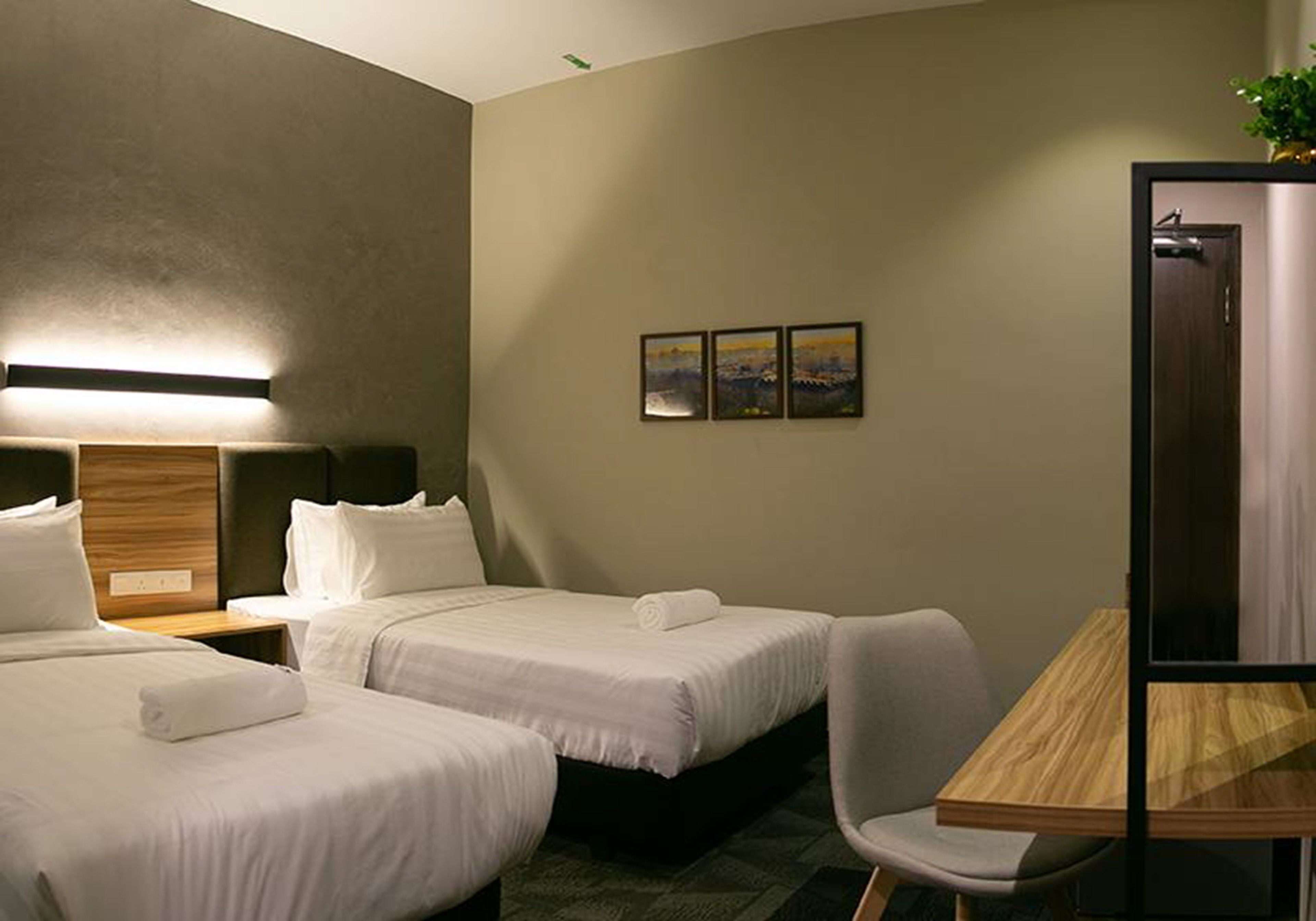 Quarto de hotel com duas camas de solteiro, uma escrivaninha, uma cadeira e uma mesinha. Há luminárias fixadas na parede e um quadro emoldurado sobre as camas.