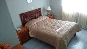 Cama bem arrumada com colcha estampada, mesa de cabeceira de madeira com luminária, telefone e pequena mesa com projetor.