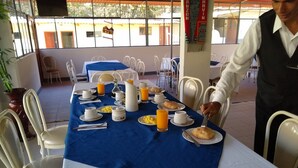 Disposição para o café da manhã, com uma mesa coberta por uma toalha azul, cadeiras brancas e itens como ovos, suco e doces.