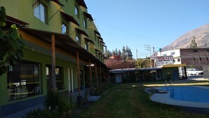 Um hotel com piscina, área externa coberta e uma montanha ao fundo.