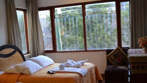 Quarto de hotel com uma cama, uma janela com vista para árvores, uma mesinha com um vaso de flores e uma cadeira com almofada colorida.