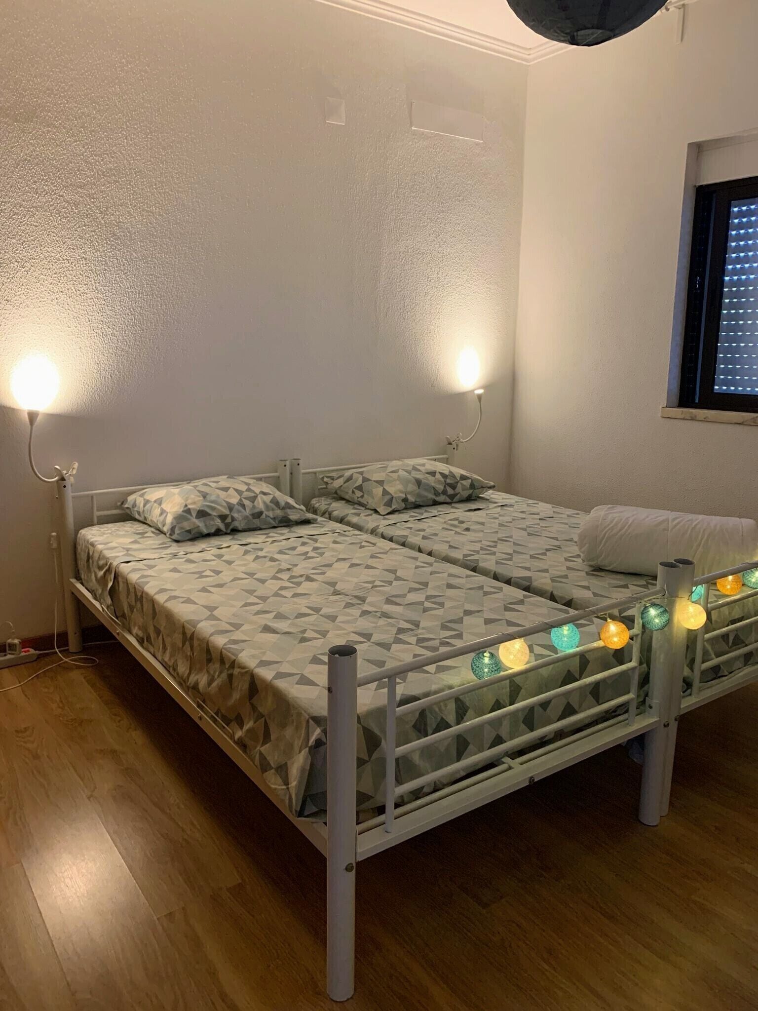 Uma cama de casal com roupa de cama estampada em padrão geométrico, duas luminárias de cabeceira e um fio de luzes coloridas.