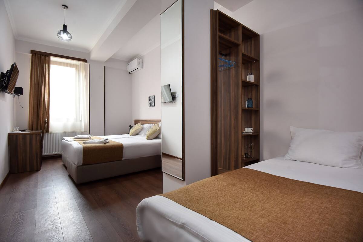 Quarto de hotel com duas camas, um guarda-roupa de madeira, um espelho, um radiador e uma janela com cortinas.