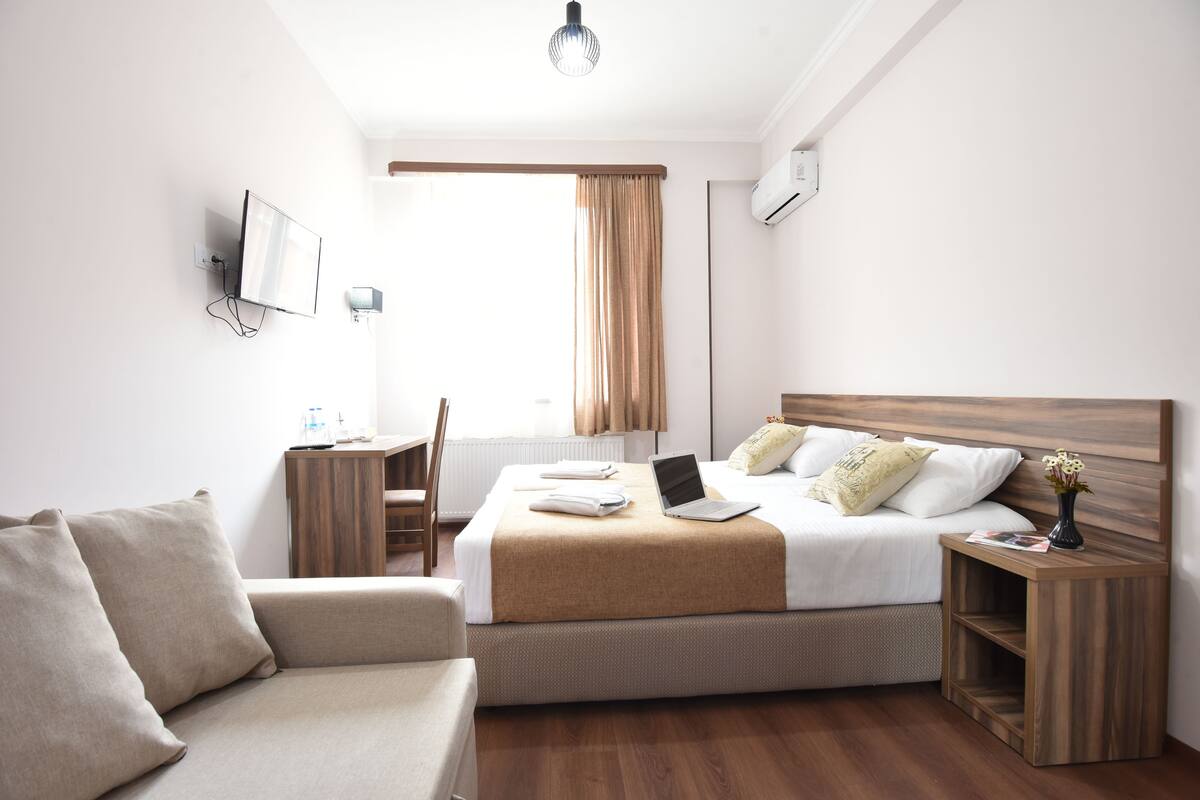 Quarto de hotel com cama, escrivaninha, cadeira, televisão e janela com cortinas.