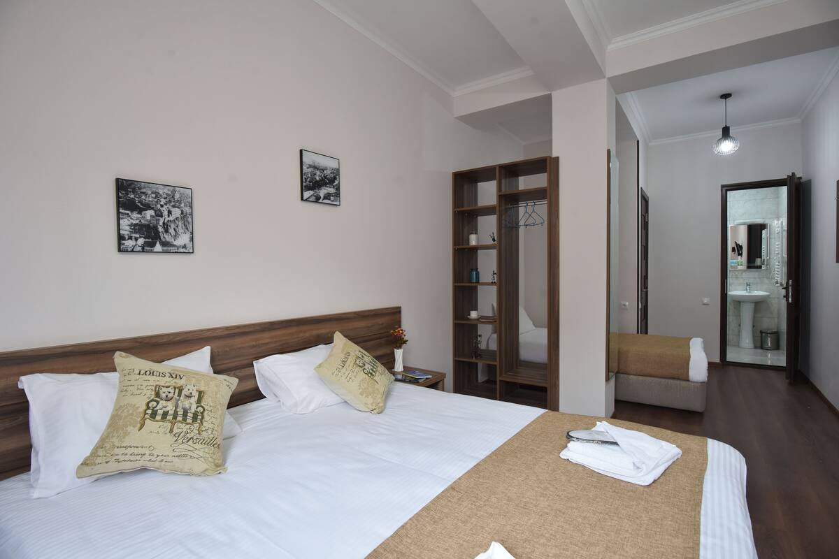 Quarto de hotel com uma cama grande, duas almofadas, cabeceira de madeira, uma mesinha com um vaso e um guarda-roupa.